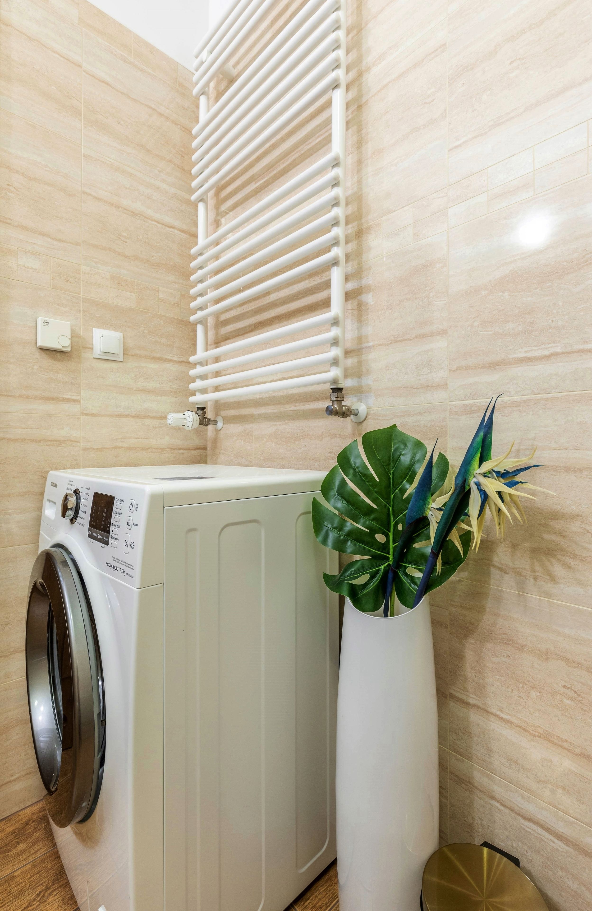 Uma lavanderia moderna com máquina de lavar de abertura frontal, um radiador branco e um vaso de planta.