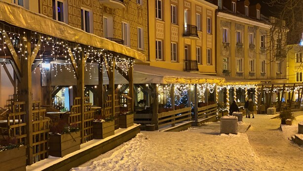 Área externa coberta de neve com bancos e mesas de madeira, cercada por edifícios com janelas iluminadas e decorações festivas.