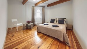 Quarto com piso de madeira, cama com manta bege, cadeira branca, mesinha com um vaso e janela com cortinas.