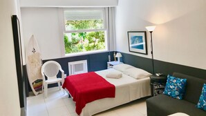 Quarto com cama, uma cadeira, uma prancha de surf, janela com vista, ventilador de teto e luminária.