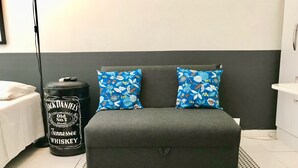 Quarto moderno com sofá cinza, duas almofadas azuis com estampas florais, um cesto de lixo preto com a etiqueta 'Jack Daniel's Tennessee Whiskey' e uma mesa de cabeceira branca.