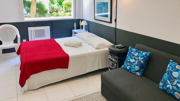Quarto de hotel com cama, sofá, poltrona, janela com vista para o verde, luminária e quadro na parede.