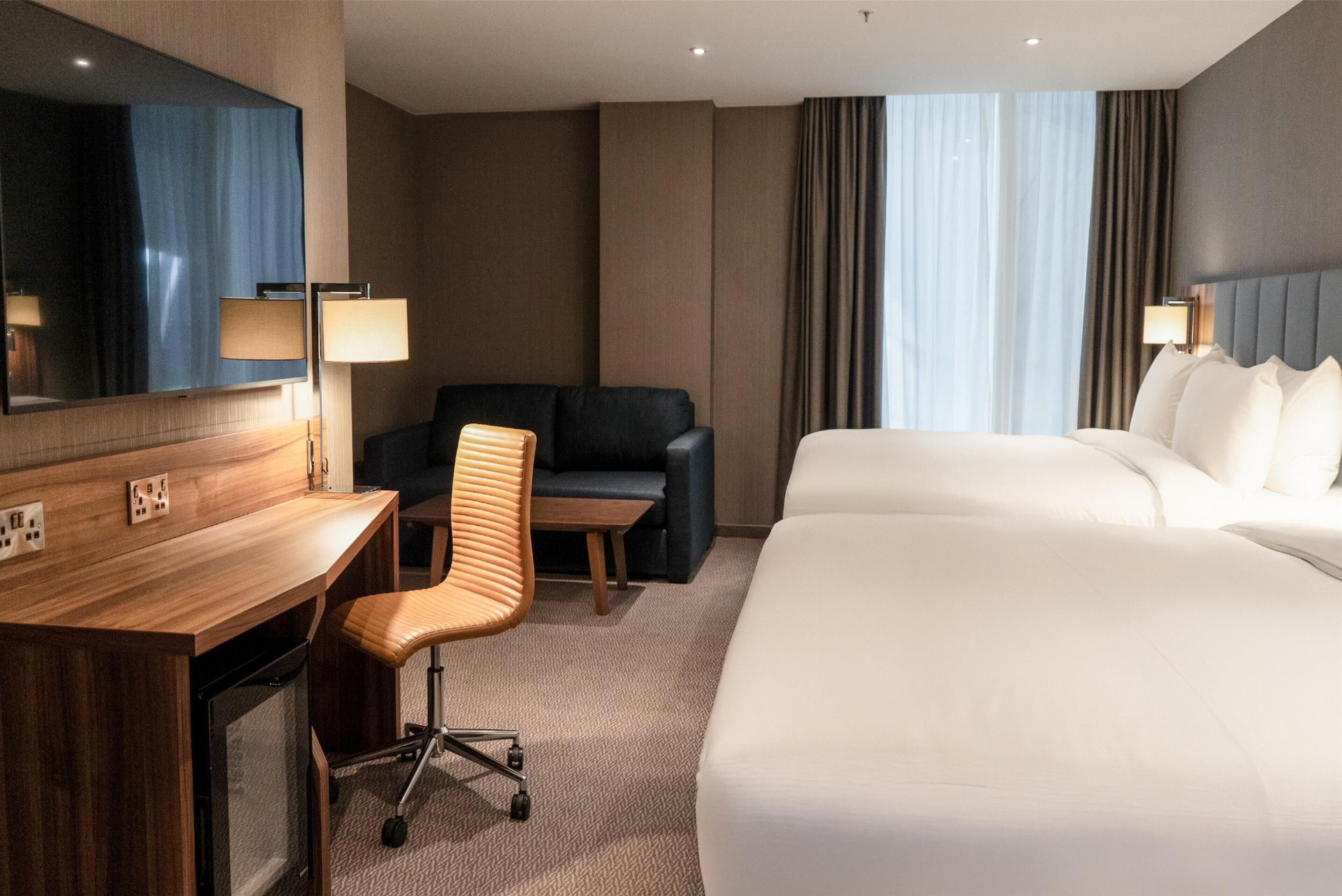 Quarto de hotel com uma cama grande, uma escrivaninha com cadeira, um sofá pequeno, uma televisão e uma janela com cortinas.