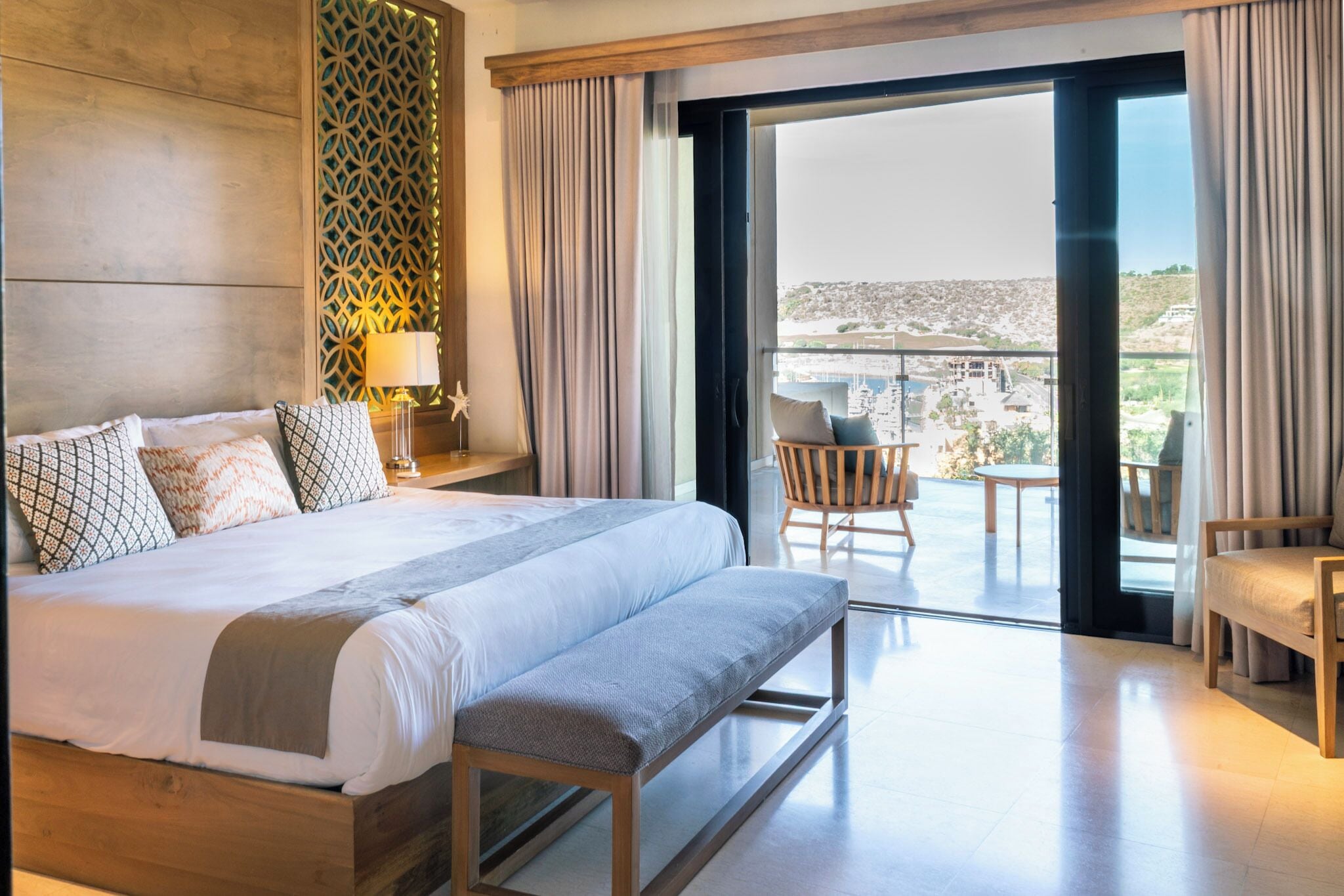 Quarto moderno com uma cama grande, um banco, uma mesa de cabeceira com abajur e vista para o exterior através de uma porta de vidro deslizante.