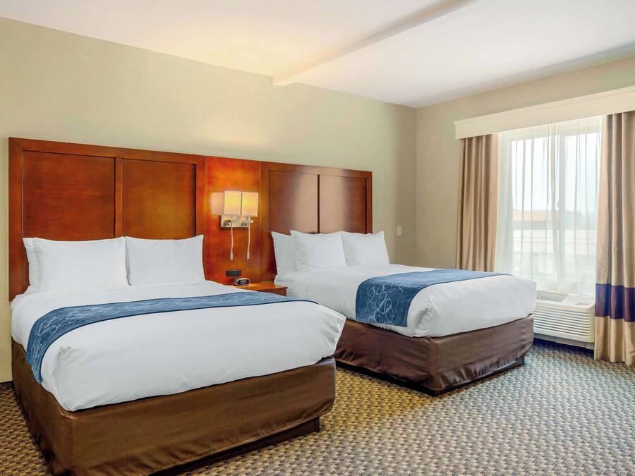 Quarto de hotel com duas camas, cabeceira de madeira, mesa de cabeceira, abajur e janela com cortinas.
