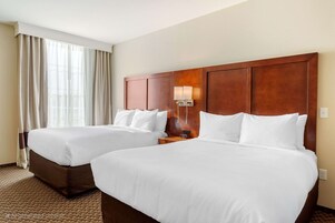 Quarto de hotel com duas camas, cabeceira de madeira, mesa de cabeceira com luminária e uma janela grande com cortinas.