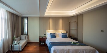 Quarto de hotel moderno com uma cama grande, mesa de cabeceira, sofá e janela com cortinas.