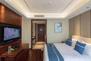 Quarto de hotel com uma cama grande, uma TV sobre um móvel de madeira e uma porta que dá para um corredor.