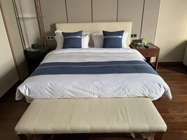 Uma cama bem arrumada com colcha branca e azul, almofada combinando e um banquinho ao pé da cama.