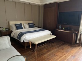 Quarto de hotel moderno com uma cama grande, piso de madeira, um banco, uma escrivaninha com televisão de tela plana e uma pequena mesa com uma garrafa de água e uma xícara.