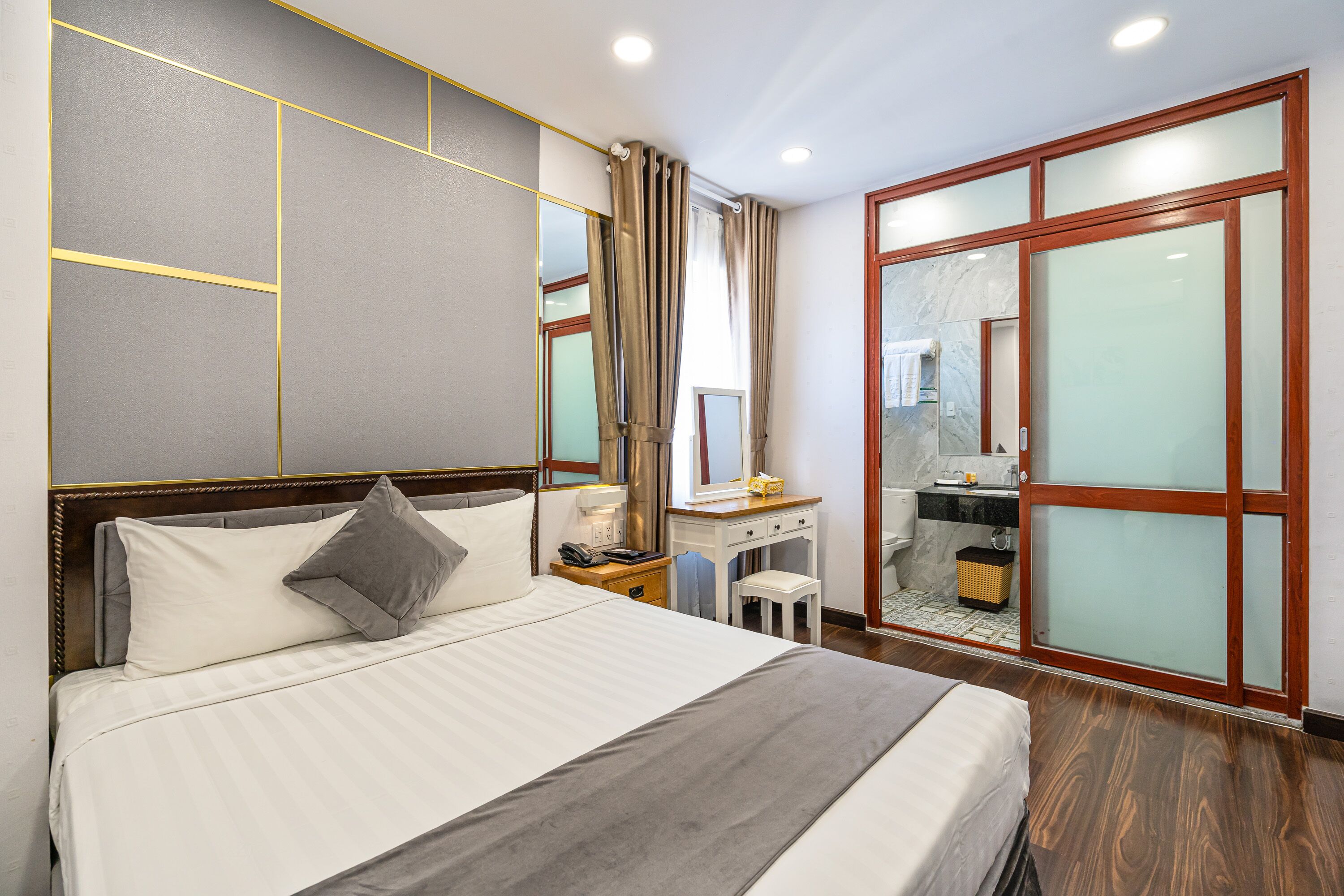 Quarto de hotel com uma cama grande, uma mesa com cadeira, toucador com espelho e porta de vidro deslizante para um banheiro.