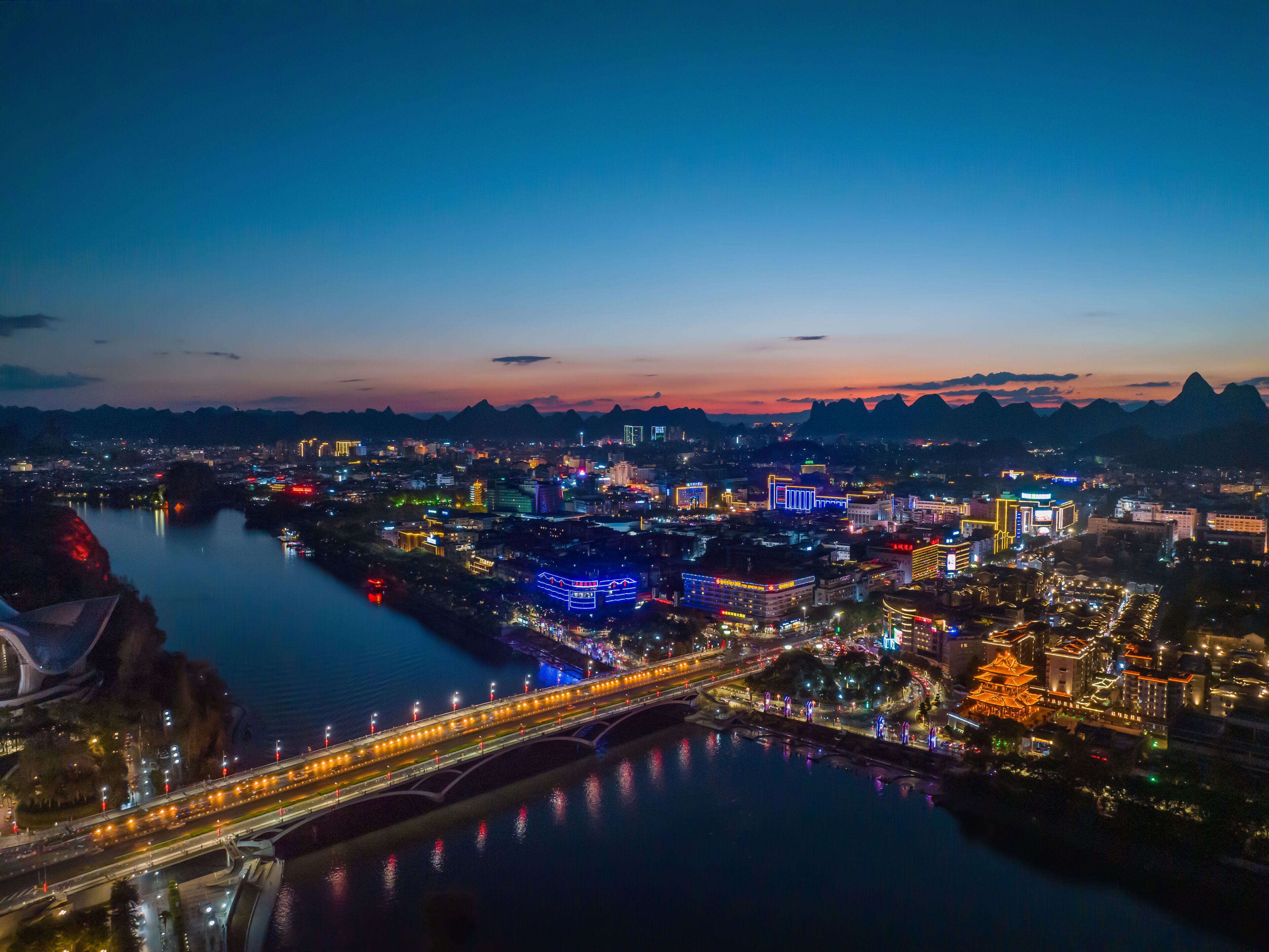 Paisagem urbana ao entardecer com edifícios iluminados, um rio e uma ponte.