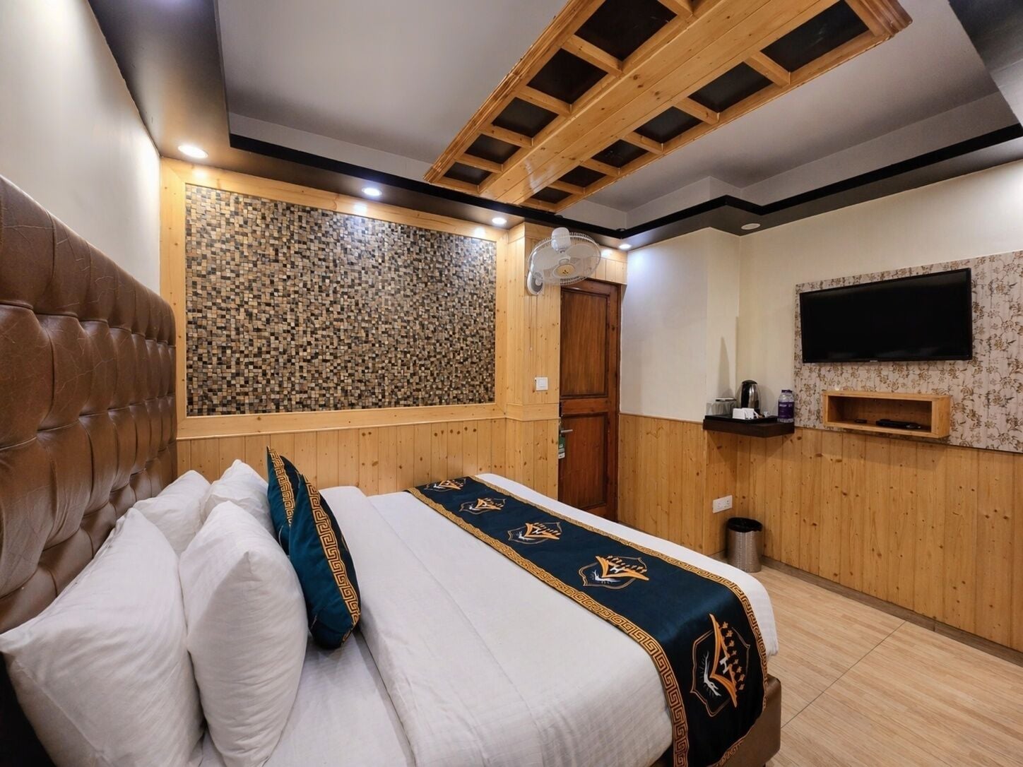 Quarto com cabeceira de madeira, cama com roupa de cama branca e azul, parede estampada, porta de madeira, uma pequena estante com objetos e uma televisão montada na parede.