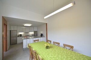 Cozinha compacta com armários brancos, geladeira de aço inoxidável e área de jantar com toalha de mesa verde e cadeiras de madeira.