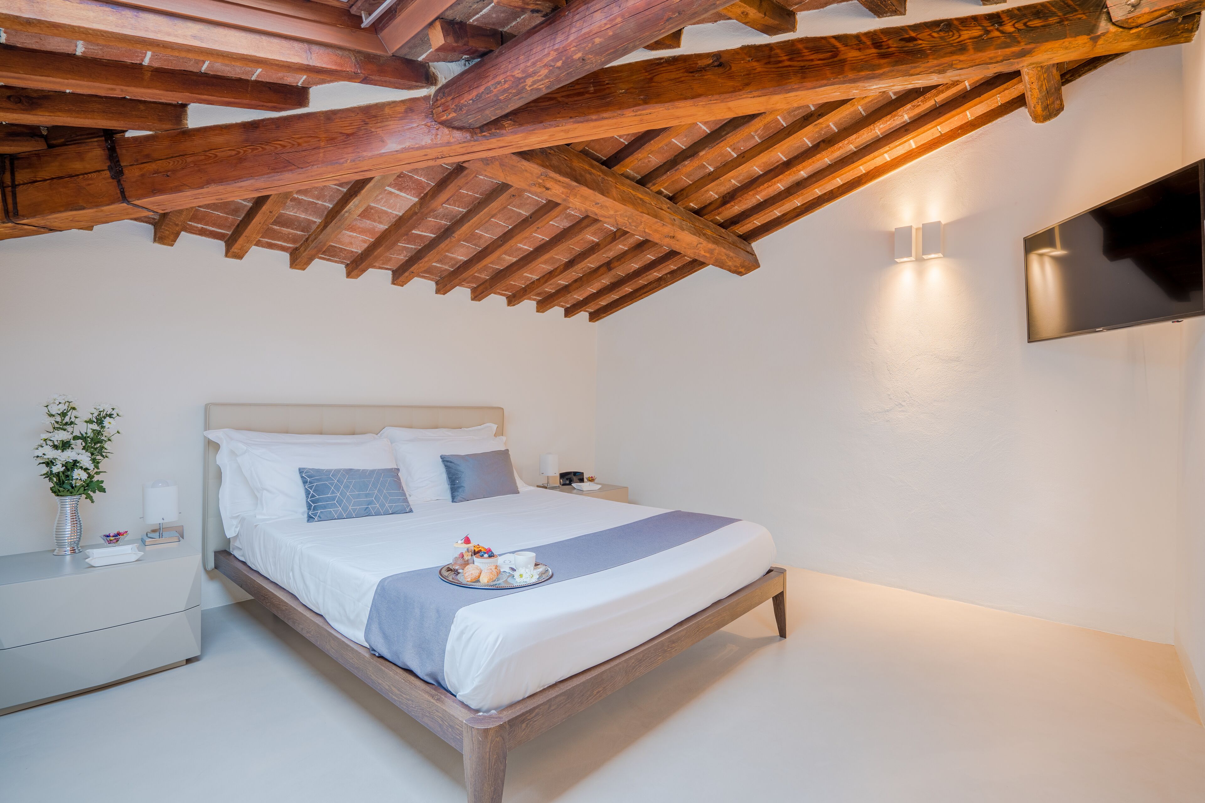 Quarto com teto de madeira, cama com roupa de cama branca, mesa de cabeceira com um vaso e televisão fixada na parede.