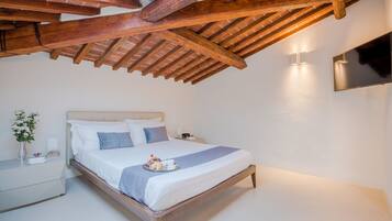 Quarto com teto de madeira, cama com roupa de cama branca, mesa de cabeceira com um vaso e televisão fixada na parede.