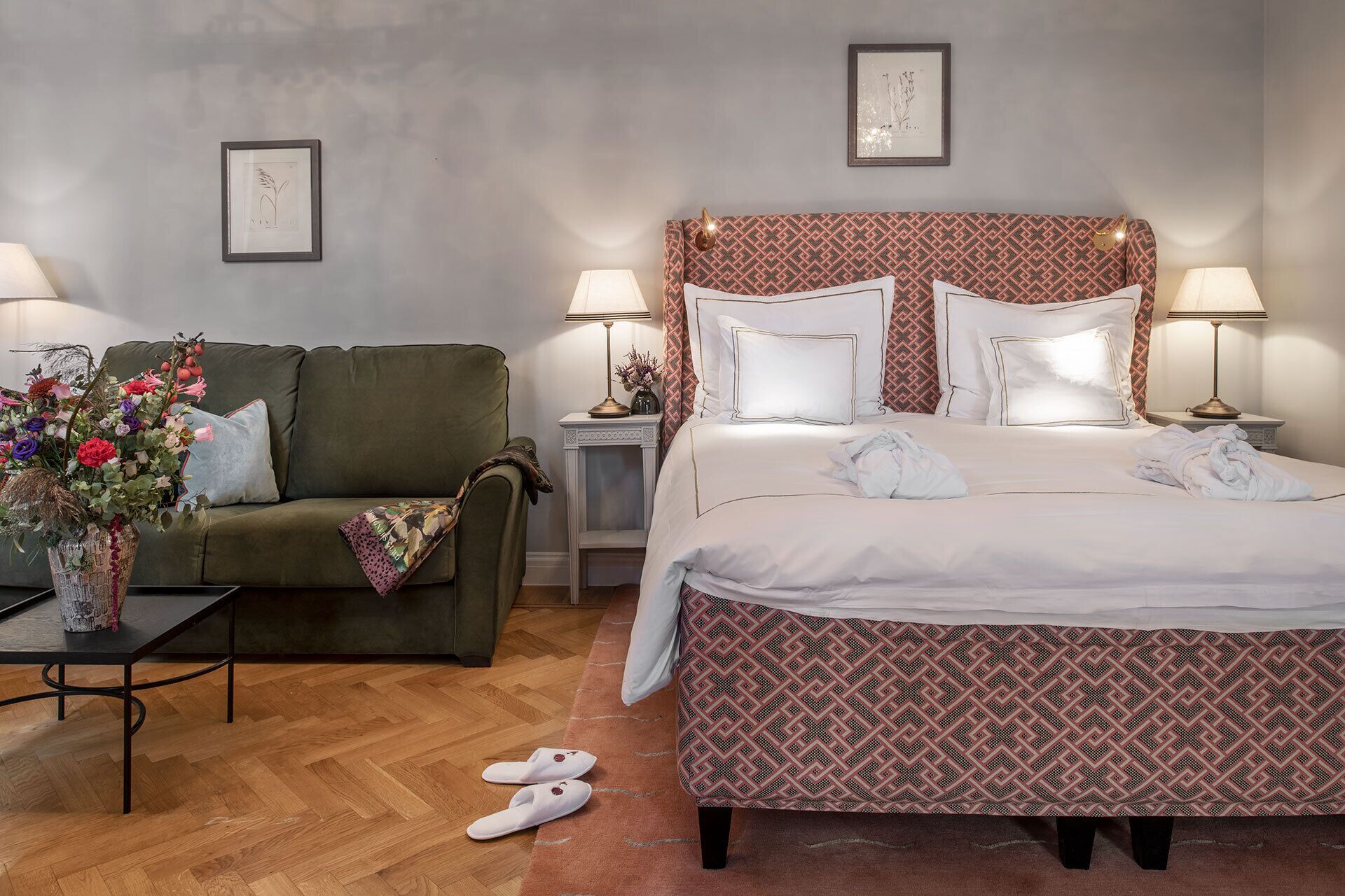 Quarto de hotel com uma cama, um sofá verde, uma mesinha com um vaso de flores e duas quadros na parede.