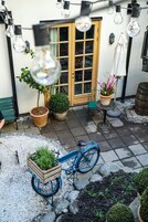 Pátio com uma bicicleta azul, plantas em vasos, uma porta de madeira e lâmpadas penduradas.