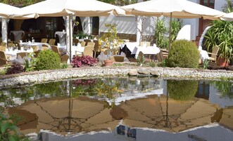 Uma área de refeições ao ar livre tranquila, com mesas e cadeiras sob grandes guarda-sóis, um pequeno lago com peixes e um jardim bem cuidado com diversas plantas.
