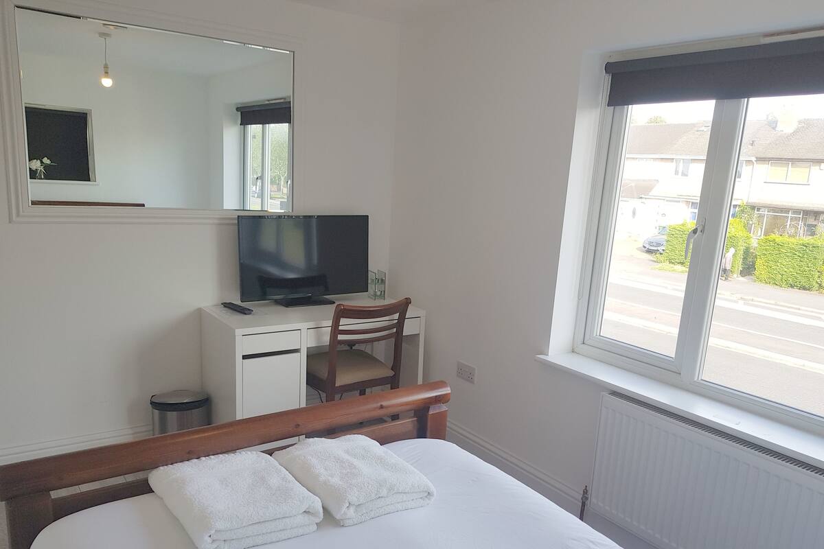 Quarto com cama, escrivaninha com computador e televisão, uma cadeira e janela com vista para a rua.