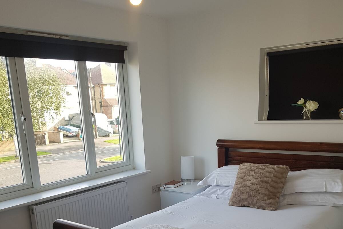 Quarto com cama de madeira, roupa de cama branca, tapete vermelho, mesa de cabeceira com luminária e janela com vista para a rua.