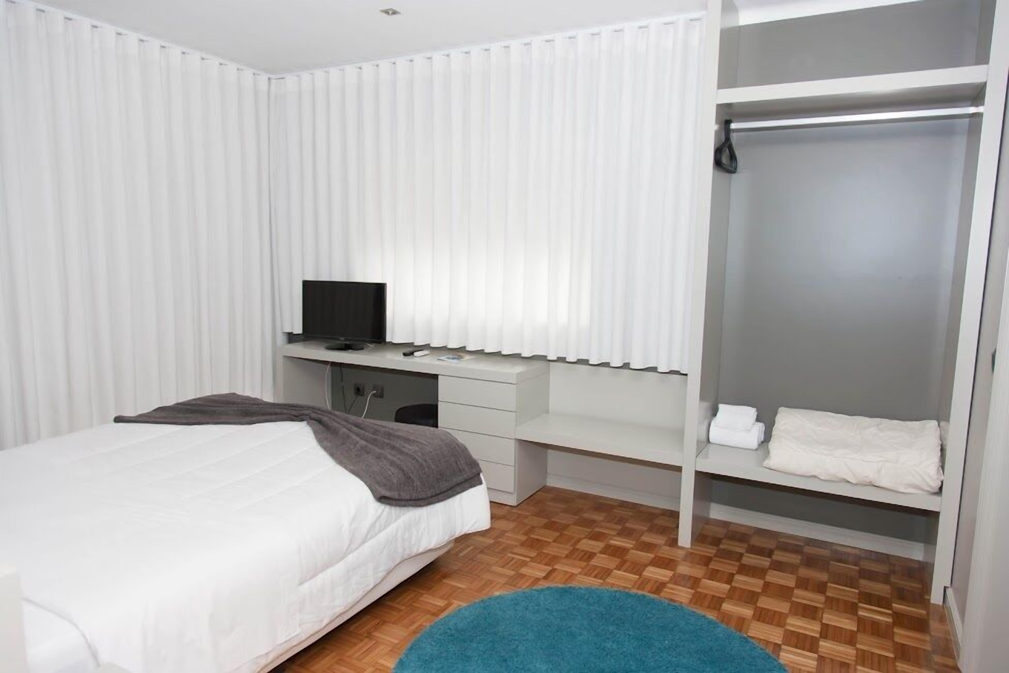 Quarto com uma cama, uma escrivaninha com televisão, guarda-roupa e um tapete azul.