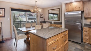 Cozinha moderna com ilha central, armários de madeira, frigorífico de aço inoxidável e área de jantar com cadeiras e mesa.