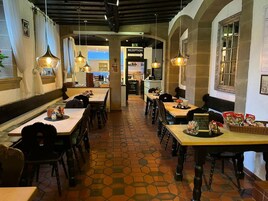 Interior de restaurante com piso em padrão hexagonal, mesas e cadeiras de madeira, iluminação pendente e uma área de recepção ao fundo.