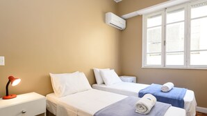 Quarto de hotel com duas camas, mesa de cabeceira com abajur, aparelho de ar condicionado e janela com persianas.