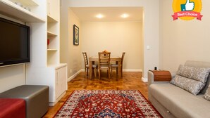 Uma sala de estar com sofá, televisão, mesa de jantar com cadeiras e tapete estampado.