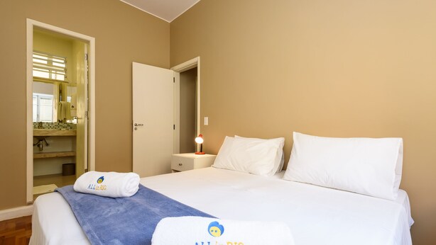 Quarto de hotel com cama, mesa de cabeceira, luminária e banheiro visível através de uma porta aberta.