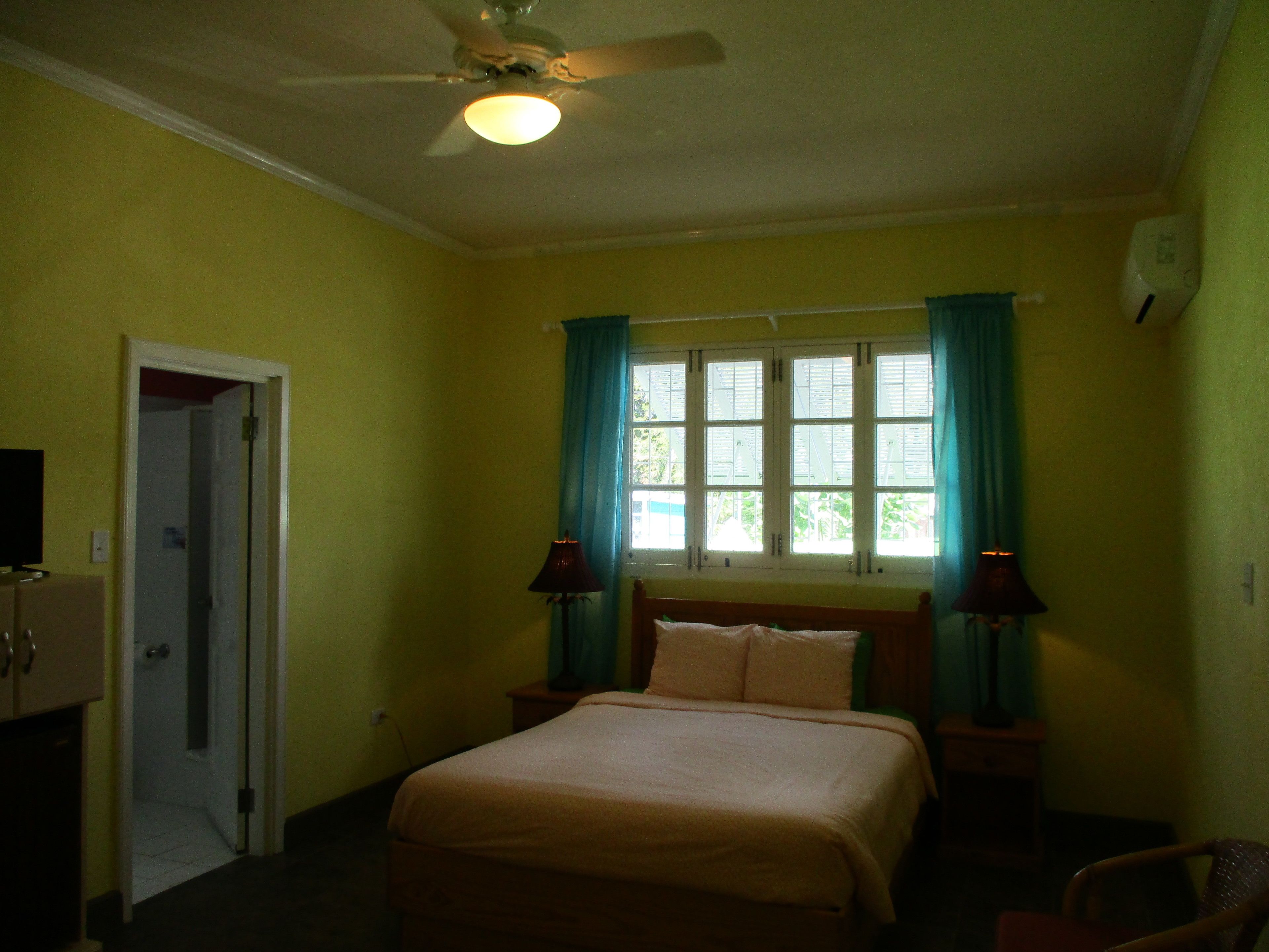 Quarto com cama, ventilador de teto, janela com cortinas e mesa de cabeceira com abajur.