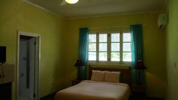 Quarto com cama, ventilador de teto, janela com cortinas e mesa de cabeceira com abajur.