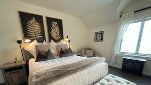 Quarto bem arrumado com uma cama grande, mesas de cabeceira, abajur, janela com cortinas e uma pequena mesa com xícaras.
