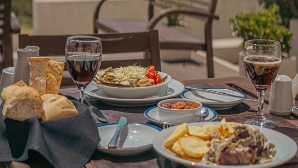Uma mesa de jantar ao ar livre com diversos pratos, copos de vinho e pão.