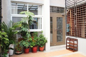 Um edifício com fachada branca, porta marrom e janela com grade de metal. Há plantas em vasos e um banco de madeira na parte externa.