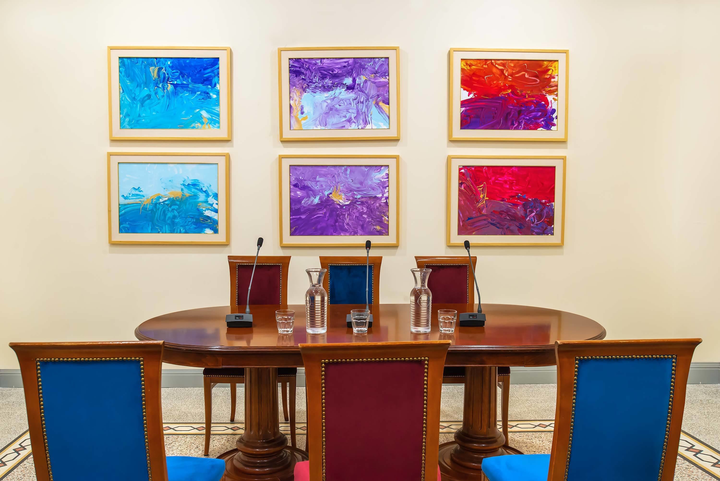 Uma sala de conferências com uma mesa redonda, seis cadeiras e seis quadros abstratos emoldurados na parede.