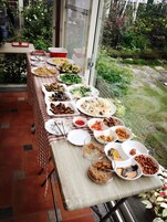 Uma mesa comprida com diversos pratos e tigelas de comida, disposta num terraço com vista para um jardim.