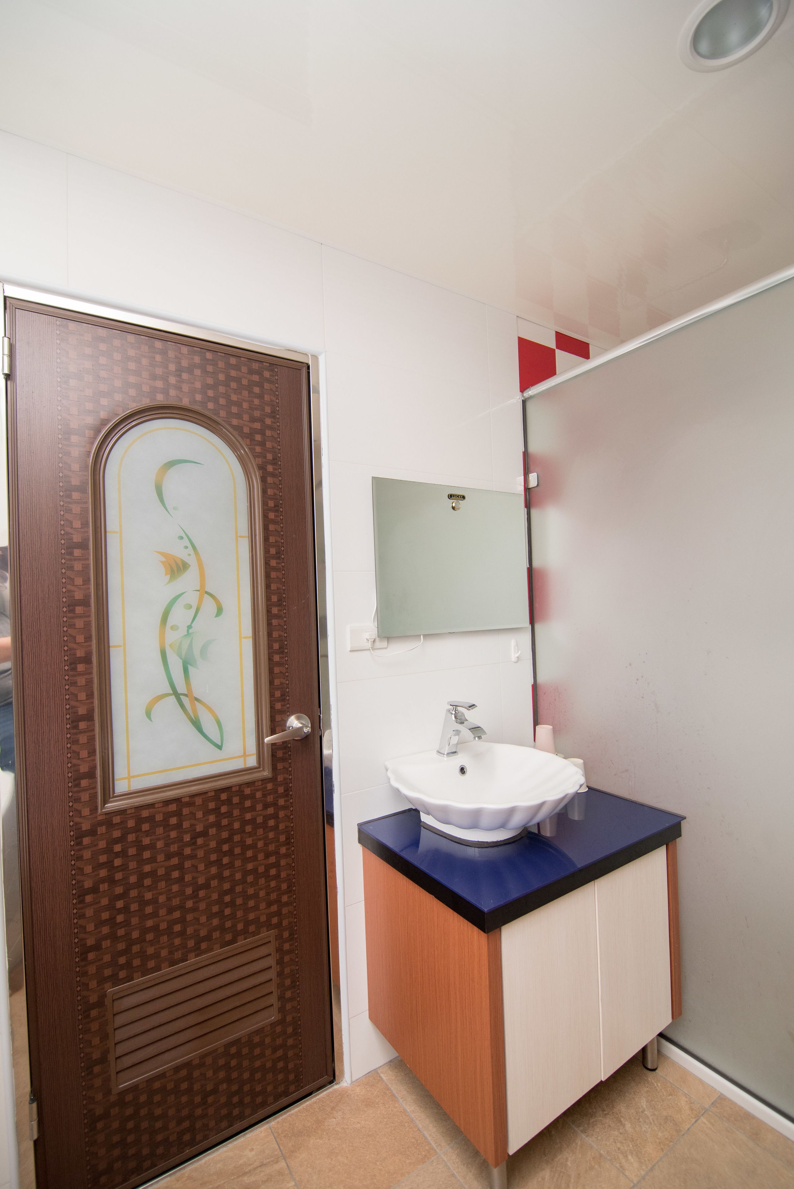 Um banheiro com porta de madeira, pia branca com bancada azul, espelho sobre a pia e piso revestido de azulejos.