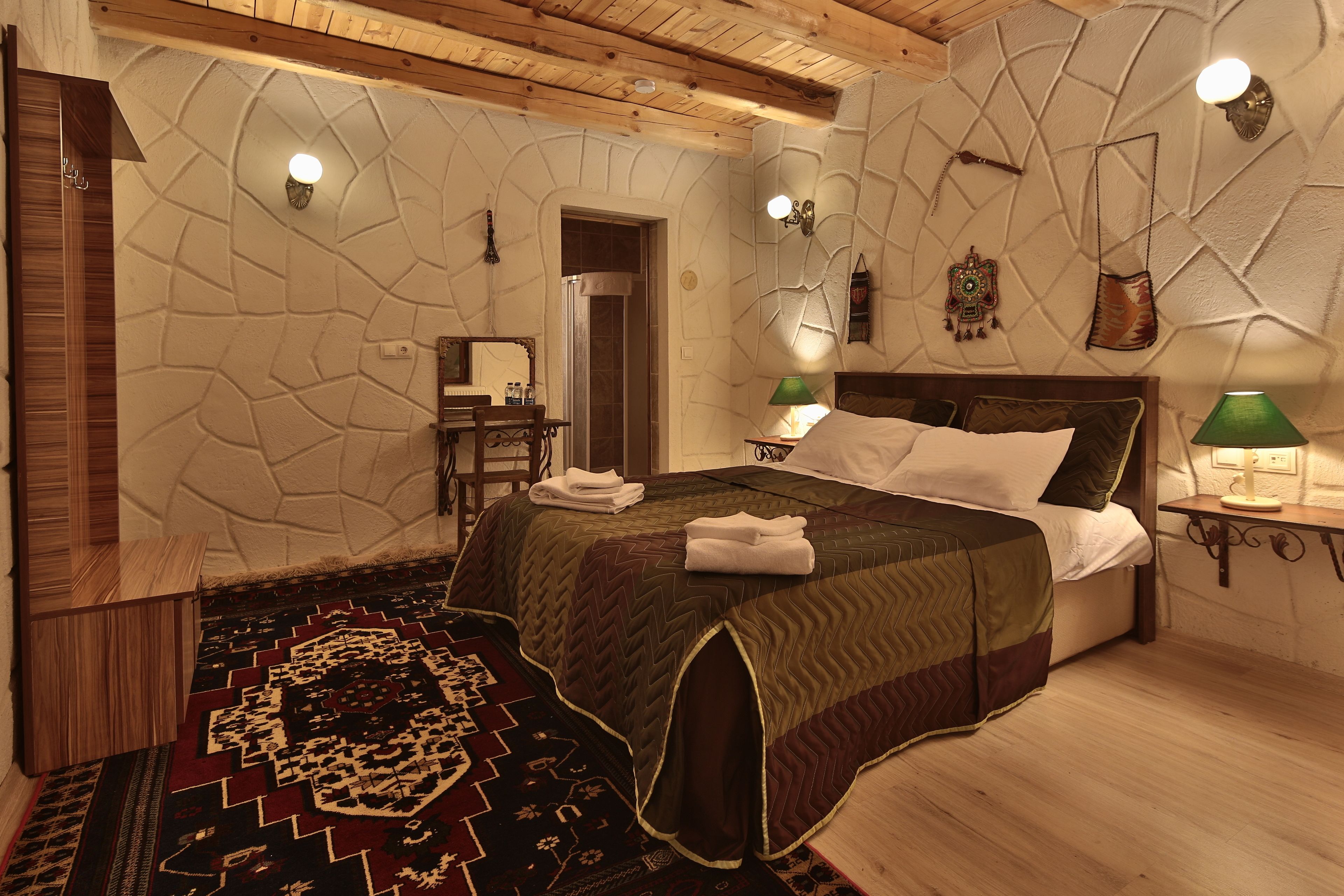 Um quarto com uma cama grande, cabeceira de madeira, mesa de cabeceira com abajur, um espelho e uma porta que dá para outro cômodo.