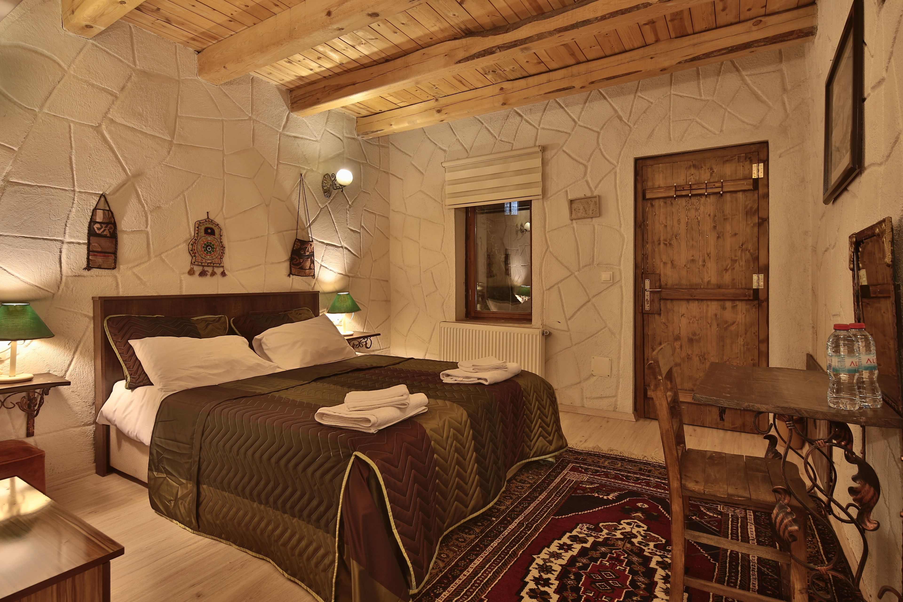 Quarto com cama, cadeira de madeira, mesa, abajur, radiador e porta.
