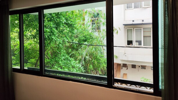 Quarto com uma grande janela que dá para uma varanda com vegetação e um edifício ao fundo.