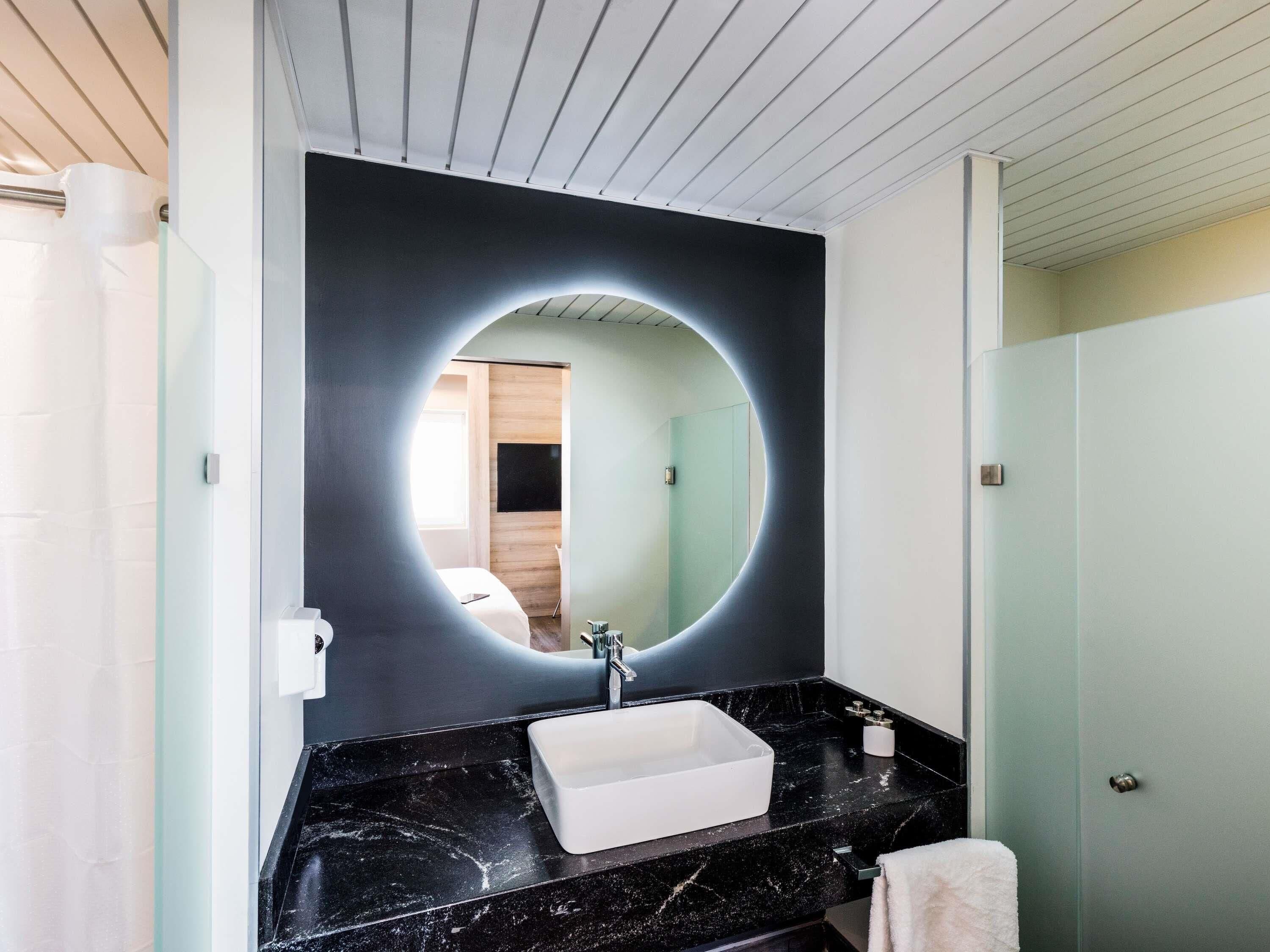 Casa de banho com lavatório de mármore preto, bacia branca e espelho grande com iluminação circular.