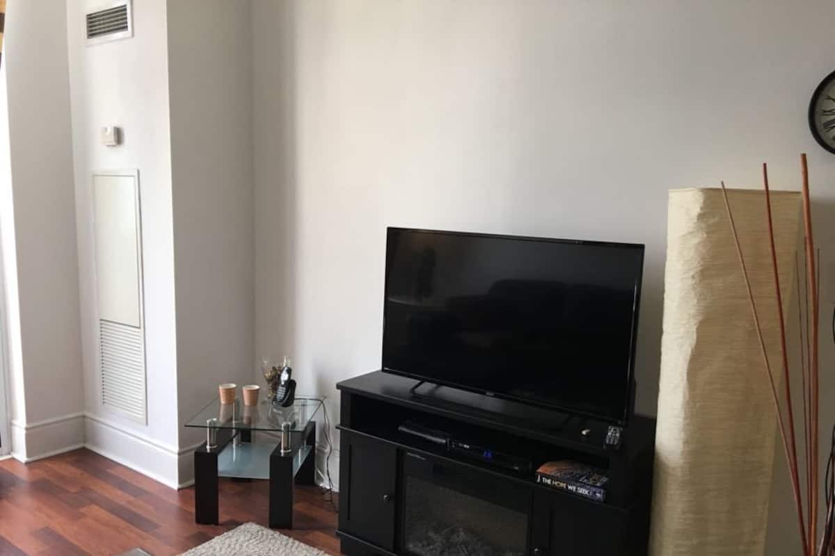 Sala de estar com TV de tela plana em suporte preto, mesa de centro de vidro, lareira e cortina bege.