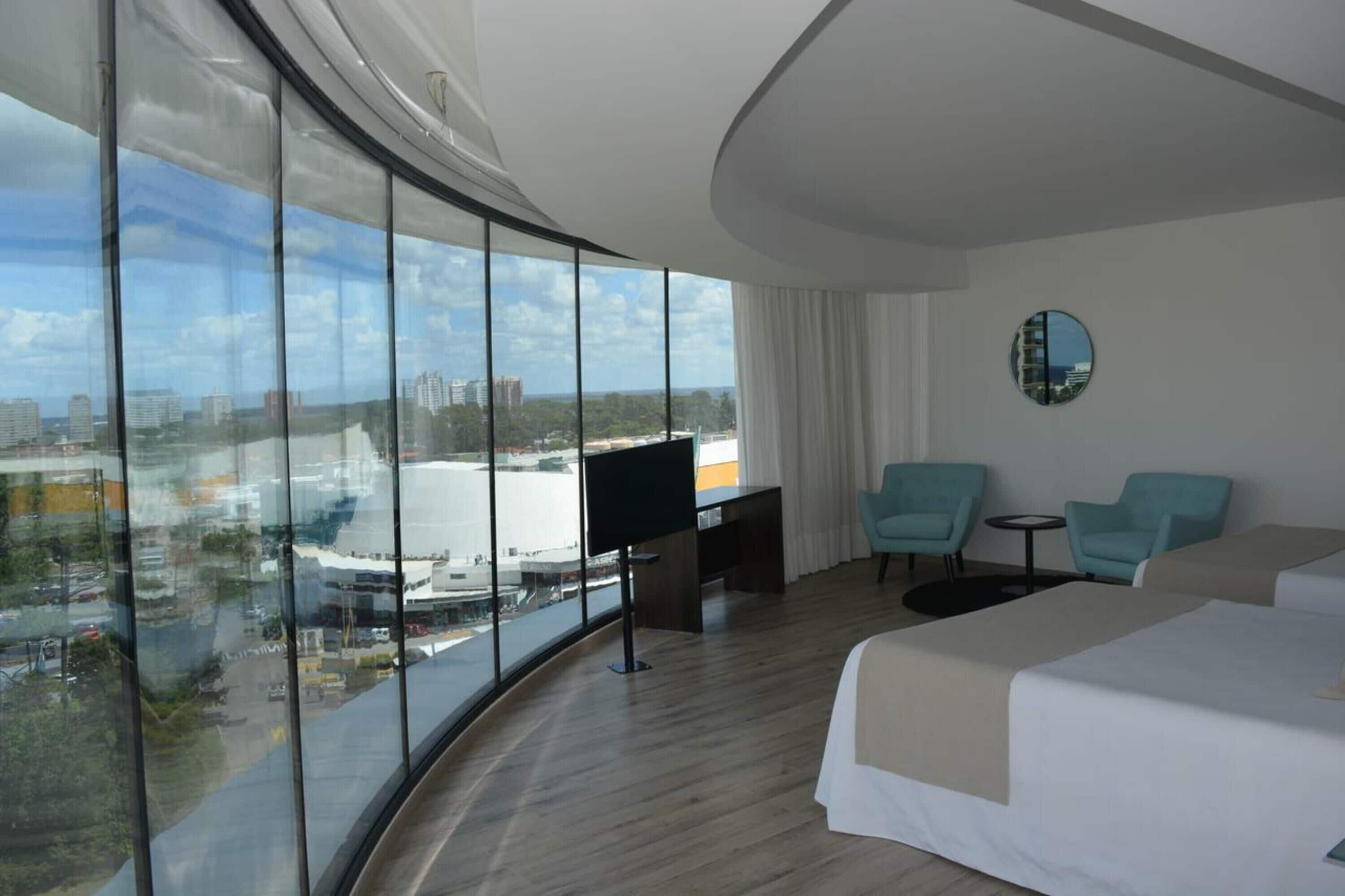 Quarto de hotel moderno com parede de vidro curva, uma cama, duas cadeiras, uma mesinha e uma televisão.