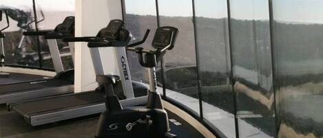 Academia com esteiras, elíptico e bicicleta ergométrica, com amplas janelas e vista para a paisagem.