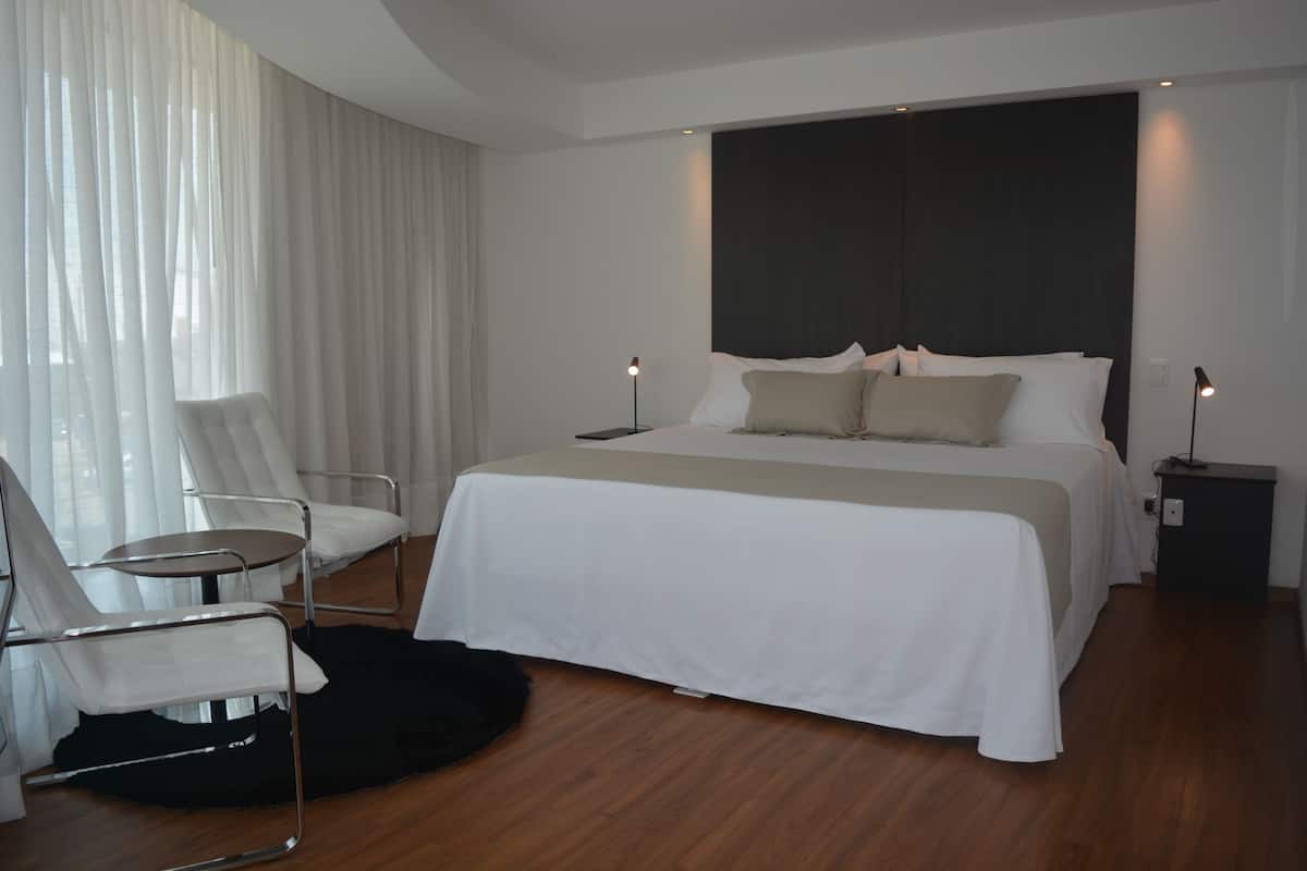 Quarto de hotel moderno com uma cama grande, uma cadeira, uma mesinha e um tapete preto.