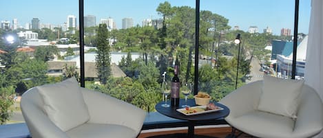 Quarto moderno com duas cadeiras brancas, uma mesinha com uma garrafa de vinho e um prato de comida. Uma grande janela oferece vista para o horizonte urbano e áreas verdes.