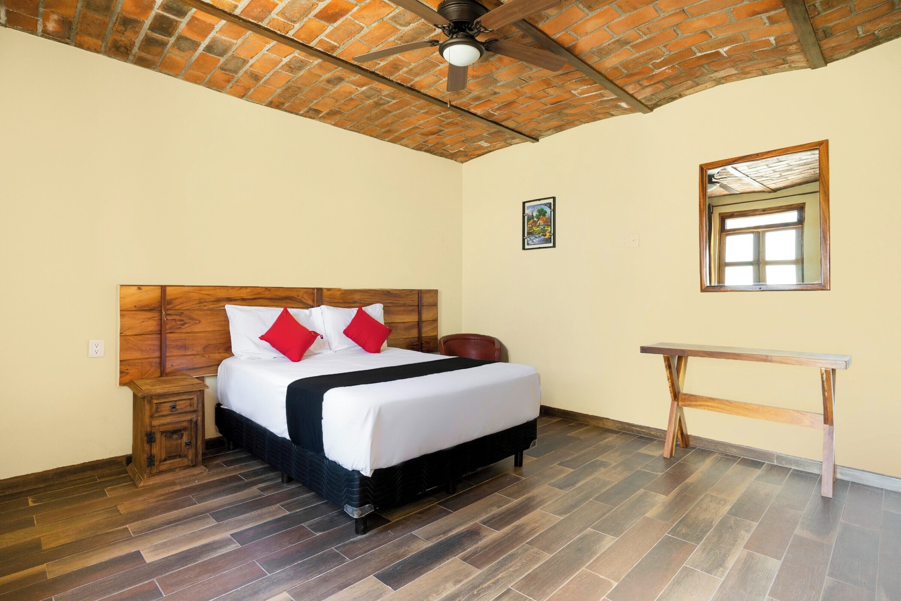 Um quarto com cabeceira de madeira, uma cama com roupa de cama branca e almofadas vermelhas, um criado-mudo de madeira, uma mesa de madeira e um ventilador de teto.