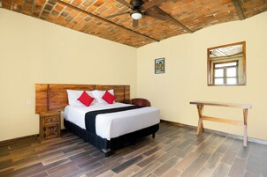 Um quarto com cabeceira de madeira, uma cama com roupa de cama branca e almofadas vermelhas, um criado-mudo de madeira, uma mesa de madeira e um ventilador de teto.